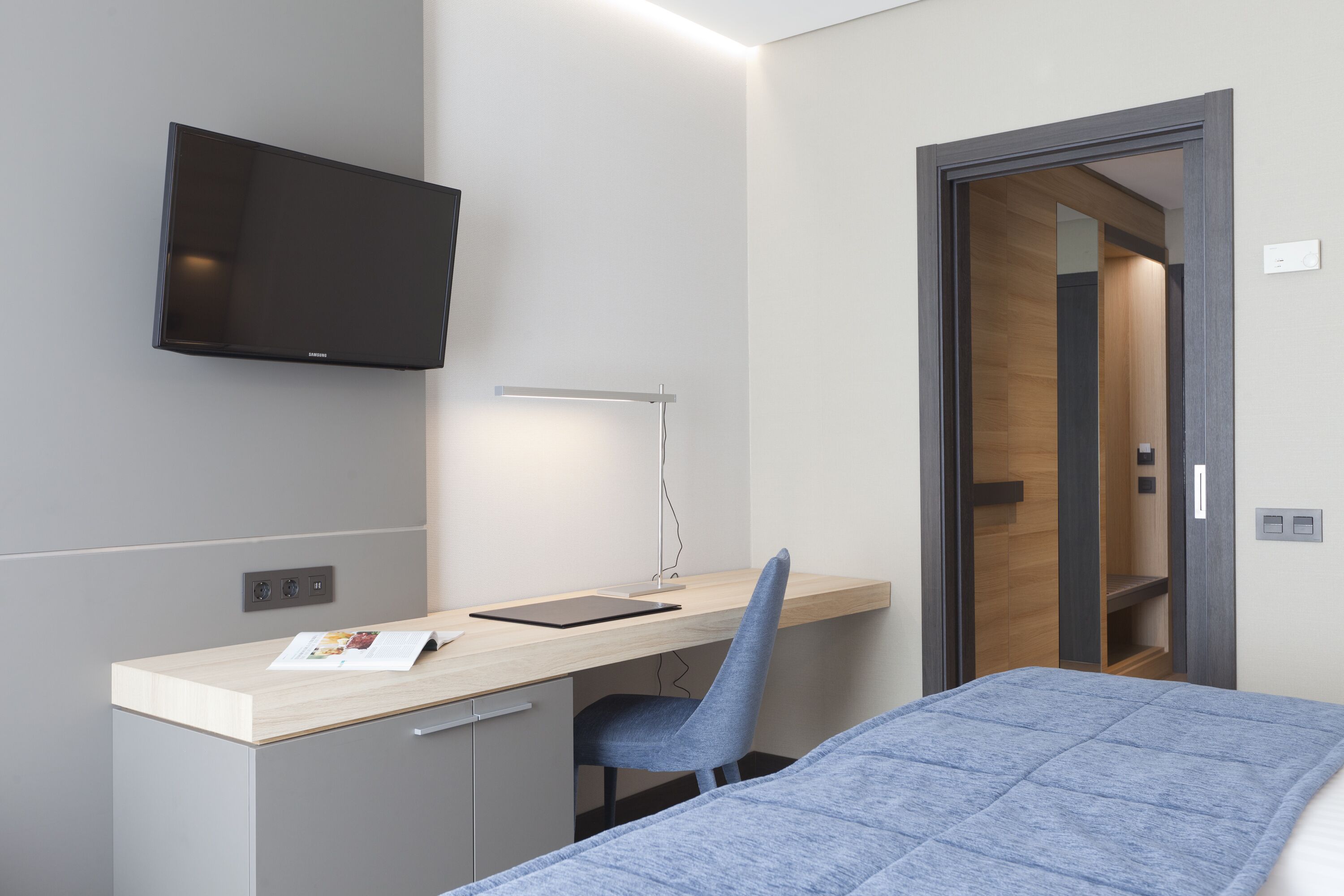 Quarto de hotel moderno com uma cama, escrivaninha com cadeira, TV de tela plana e mesa de cabeceira.