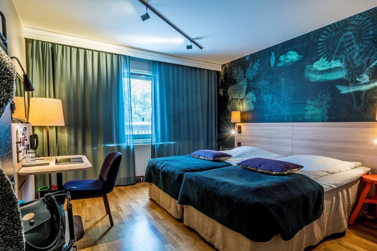 Quarto de hotel com cama, escrivaninha, cadeira e um grande mural na parede.