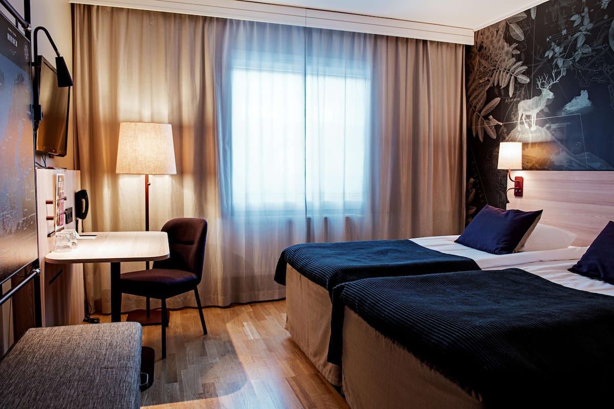 Quarto de hotel com duas camas, uma mesa com cadeira, uma luminária e uma janela grande com cortinas.