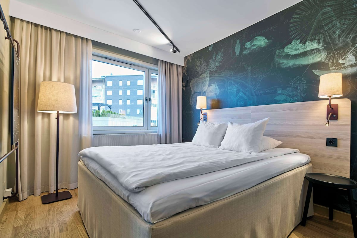 Quarto de hotel com uma cama grande, duas luminárias de cabeceira, uma janela com cortinas e um mural na parede.