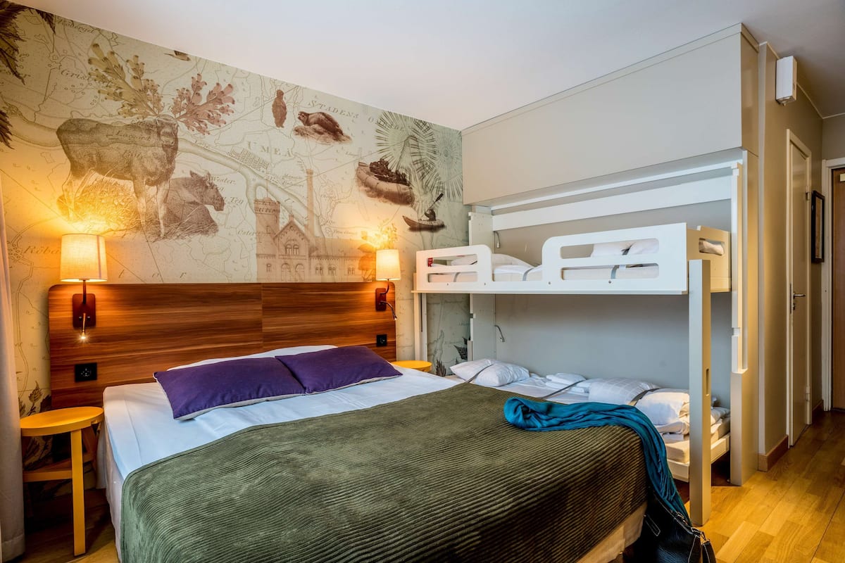 Quarto de hotel com beliche, parede decorada com tema de mapa, cabeceira de madeira e uma mesinha.