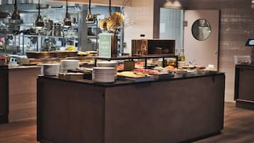 Disposição de buffet com diversos alimentos e pratos, um relógio na parede e luminárias pendentes.