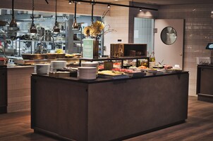 Disposição de buffet com diversos alimentos e pratos, um relógio na parede e luminárias pendentes.