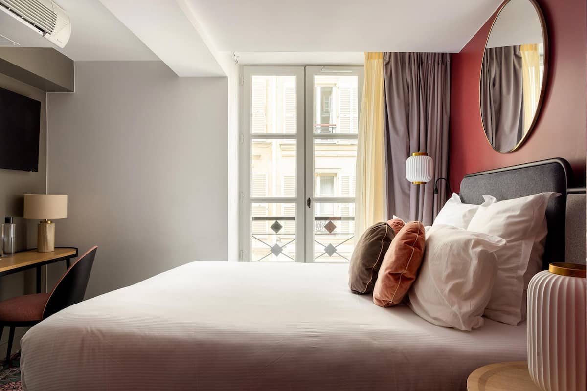 Quarto de hotel com uma cama, escrivaninha, cadeira, luminária e uma janela grande com cortinas.