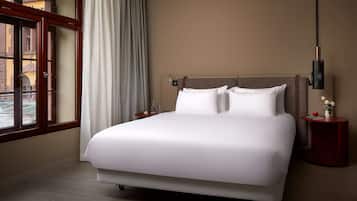 Quarto de hotel com uma cama grande, uma mesa de cabeceira com abajur e vista para prédios pela janela.