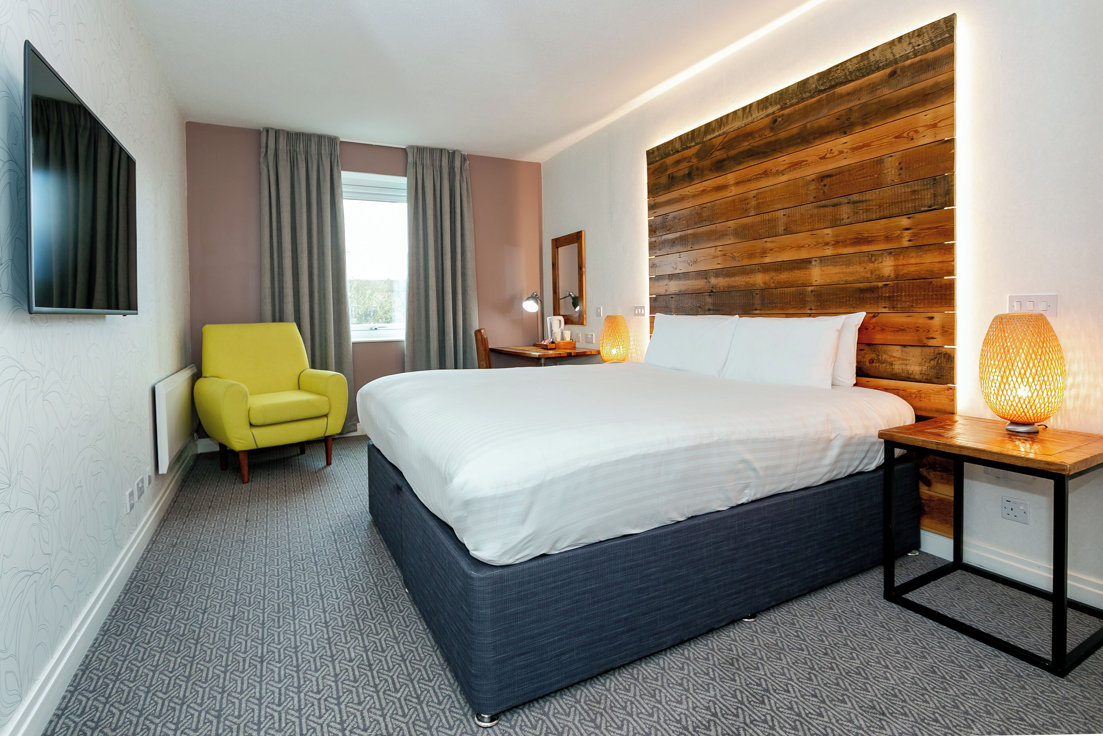Quarto de hotel com uma cama grande, uma cadeira amarela, um painel de cabeceira de madeira, uma mesa de cabeceira com abajur e uma janela com cortinas.