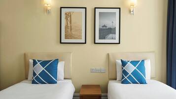 Quarto de hotel com duas camas, cada uma com travesseiro e mesa de cabeceira. Há duas quadros nas paredes, e o quarto tem uma paleta de cores neutras.