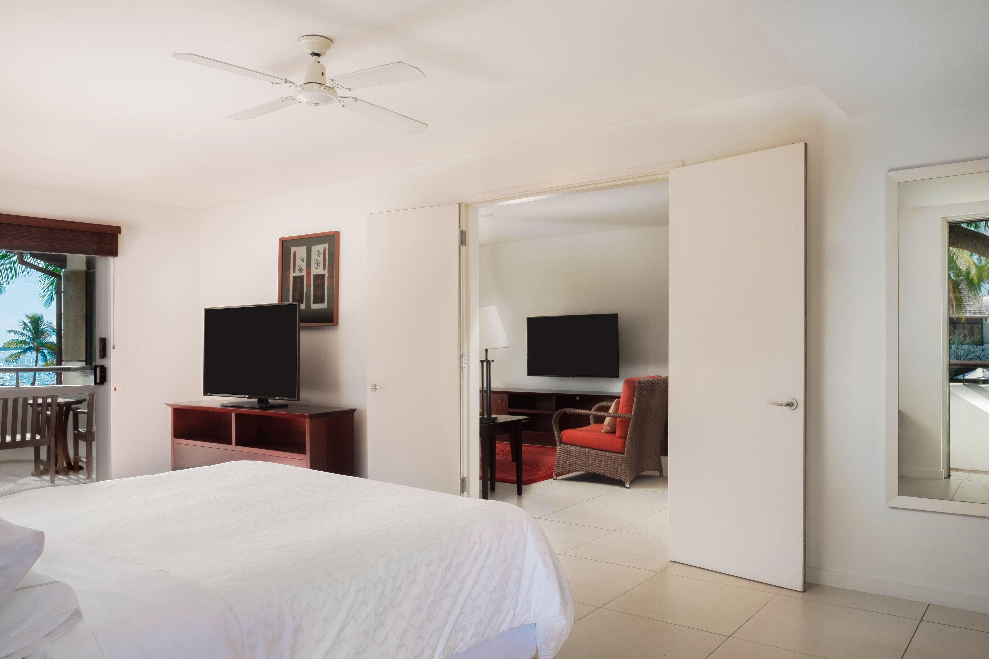 Quarto de hotel com cama, escrivaninha com televisão, uma cadeira e porta que dá para outro cômodo.