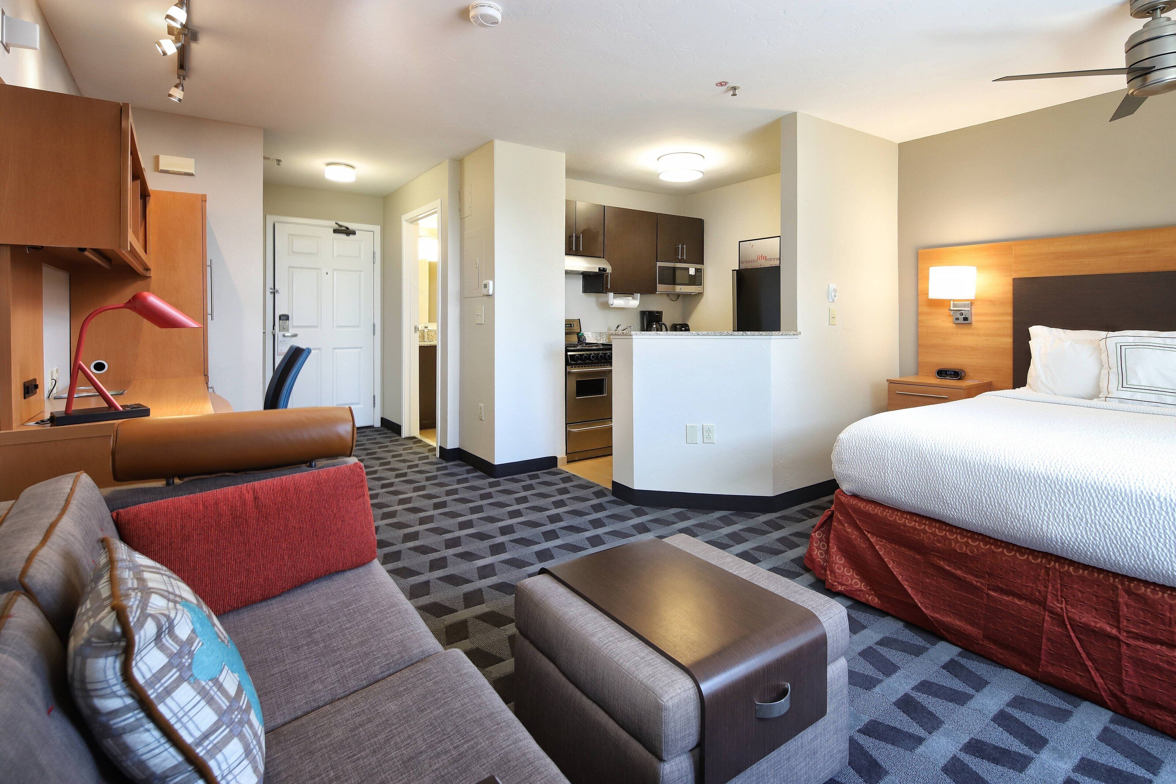 Quarto de hotel com cama, sofá, escrivaninha, luminária, cozinha compacta e banheiro.