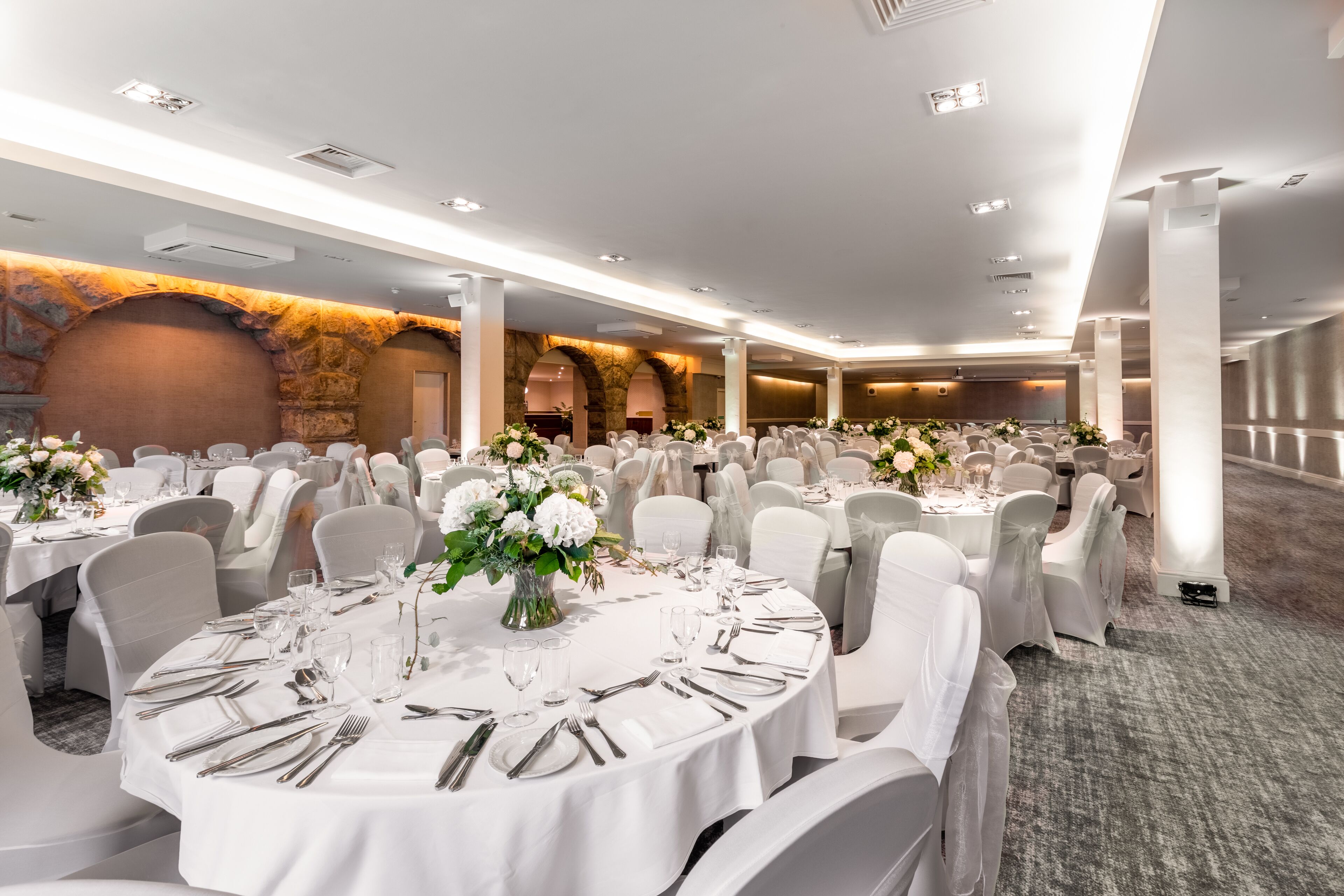 Uma sala de banquetes com mesas redondas preparadas para refeições, cadeiras brancas e centros de mesa florais.