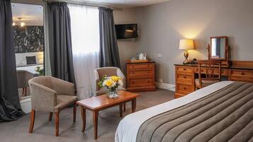 Quarto de hotel com uma cama, duas poltronas, cômoda de madeira, televisão na parede e janela com cortinas.
