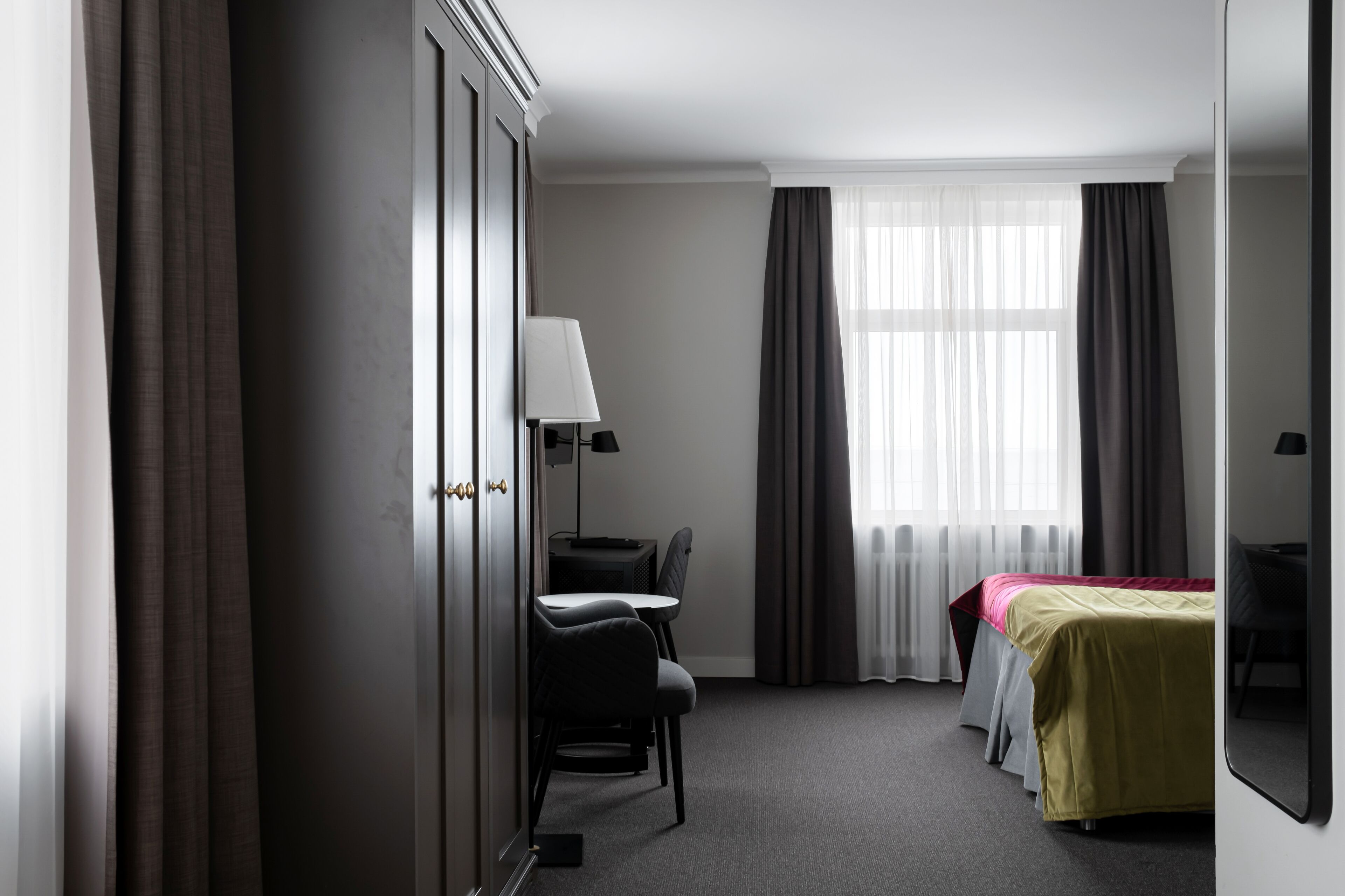 Quarto de hotel com cama, escrivaninha, cadeira, luminária e guarda-roupa.