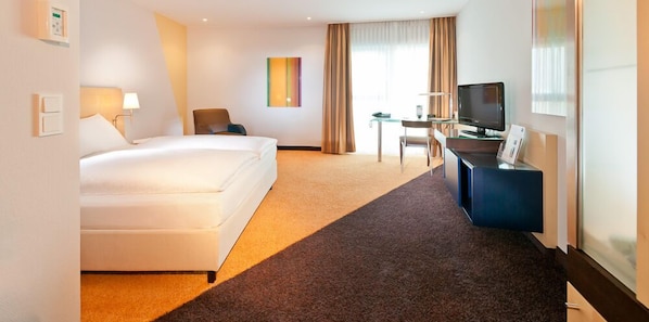 Quarto de hotel com uma cama, escrivaninha, cadeira, televisão e janela com cortinas.