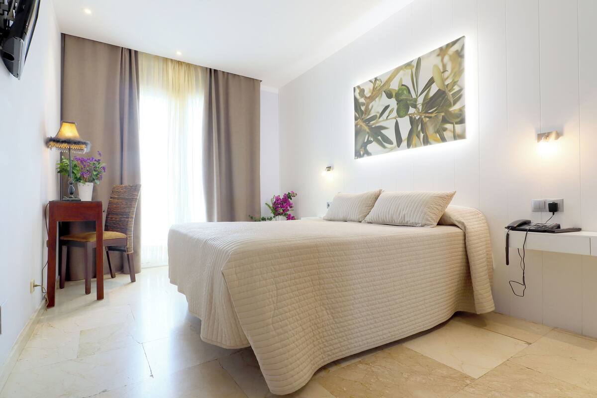 Quarto de hotel com cama, escrivaninha com luminária, cadeira, quadro na parede e janela com cortinas.