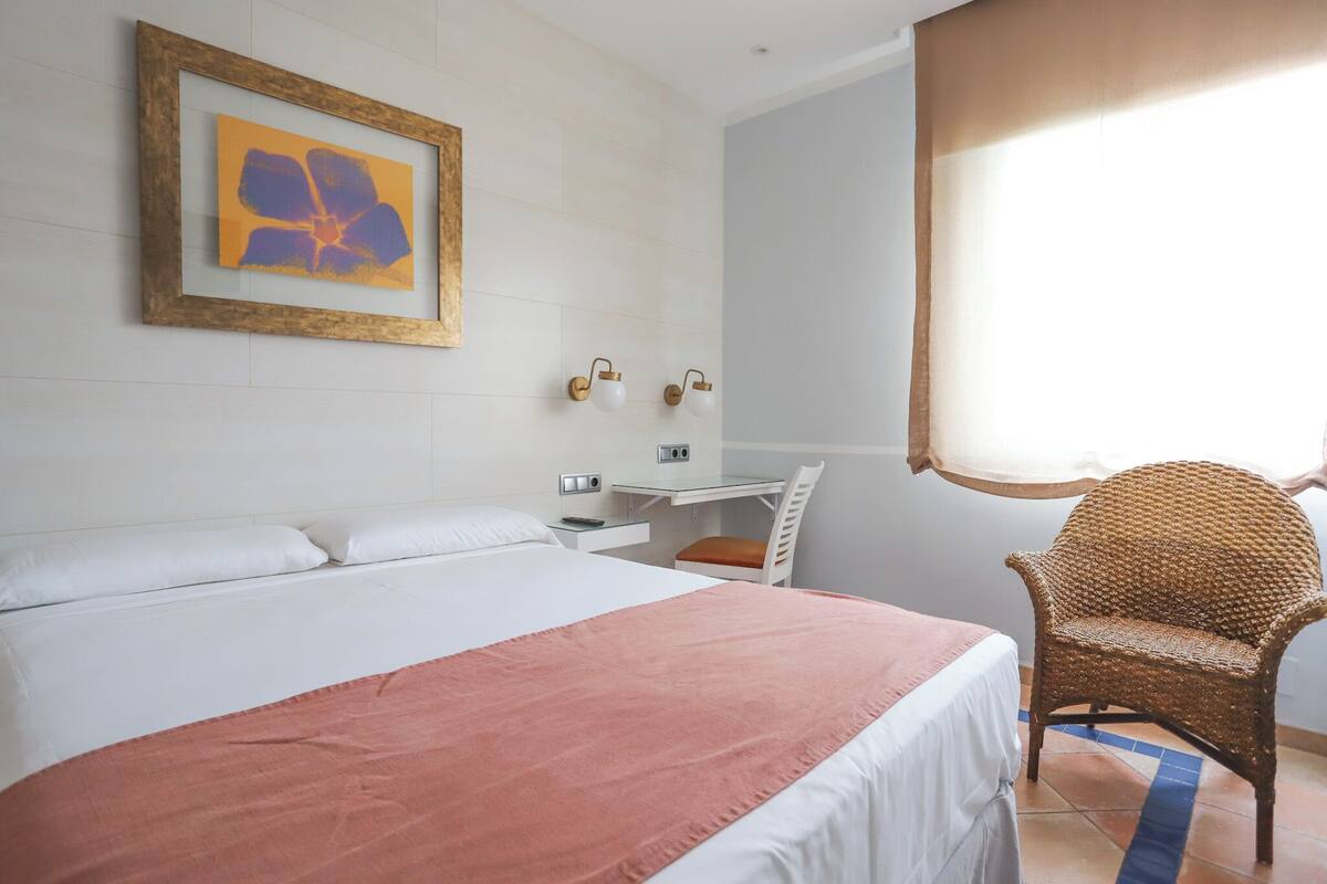 Quarto de hotel com cama, escrivaninha, cadeira, uma obra de arte emoldurada e janela com cortina.