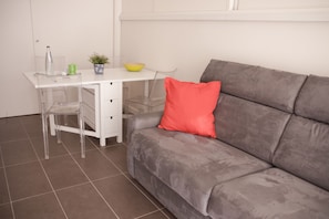 Um espaço de estar moderno com um sofá cinzento, uma mesa de vidro transparente e uma escrivaninha branca.