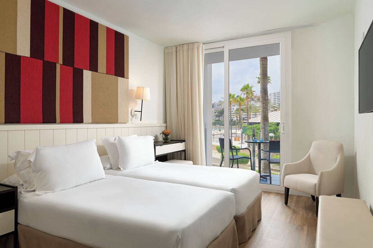 Quarto de hotel com duas camas, uma mesa, uma cadeira e vista para palmeiras e edifícios.