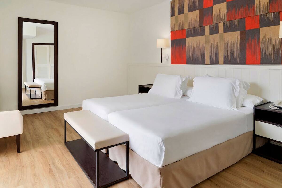 Uma cama de casal com um banco, uma mesa de cabeceira e uma luminária de parede.