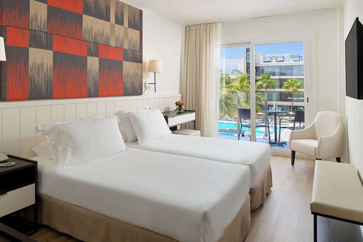 Quarto de hotel com duas camas, uma escrivaninha com luminária, uma cadeira e vista para uma piscina e palmeiras.