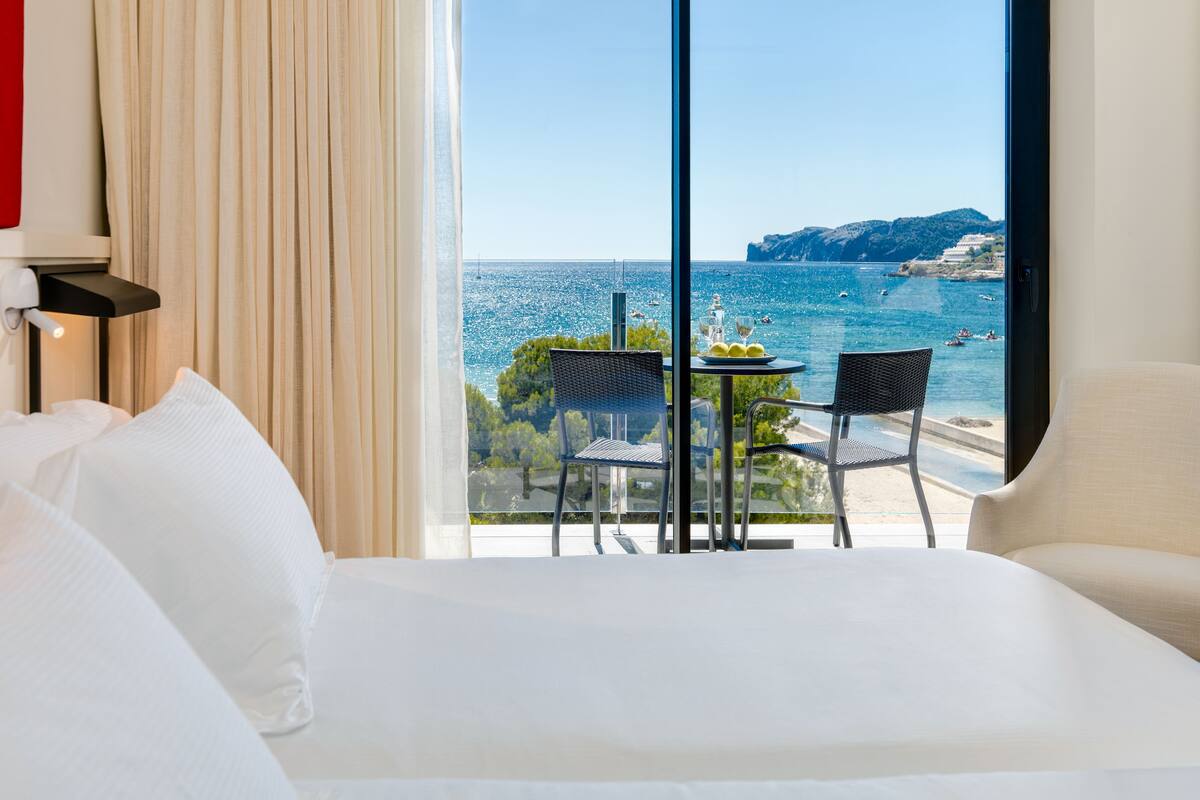 Quarto de hotel com cama, mesa e cadeiras, e vista para o mar e falésias.