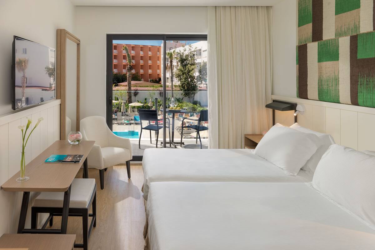 Quarto de hotel com cama, escrivaninha com cadeira, mesinha com um vaso e vista para uma piscina e edifícios lá fora.