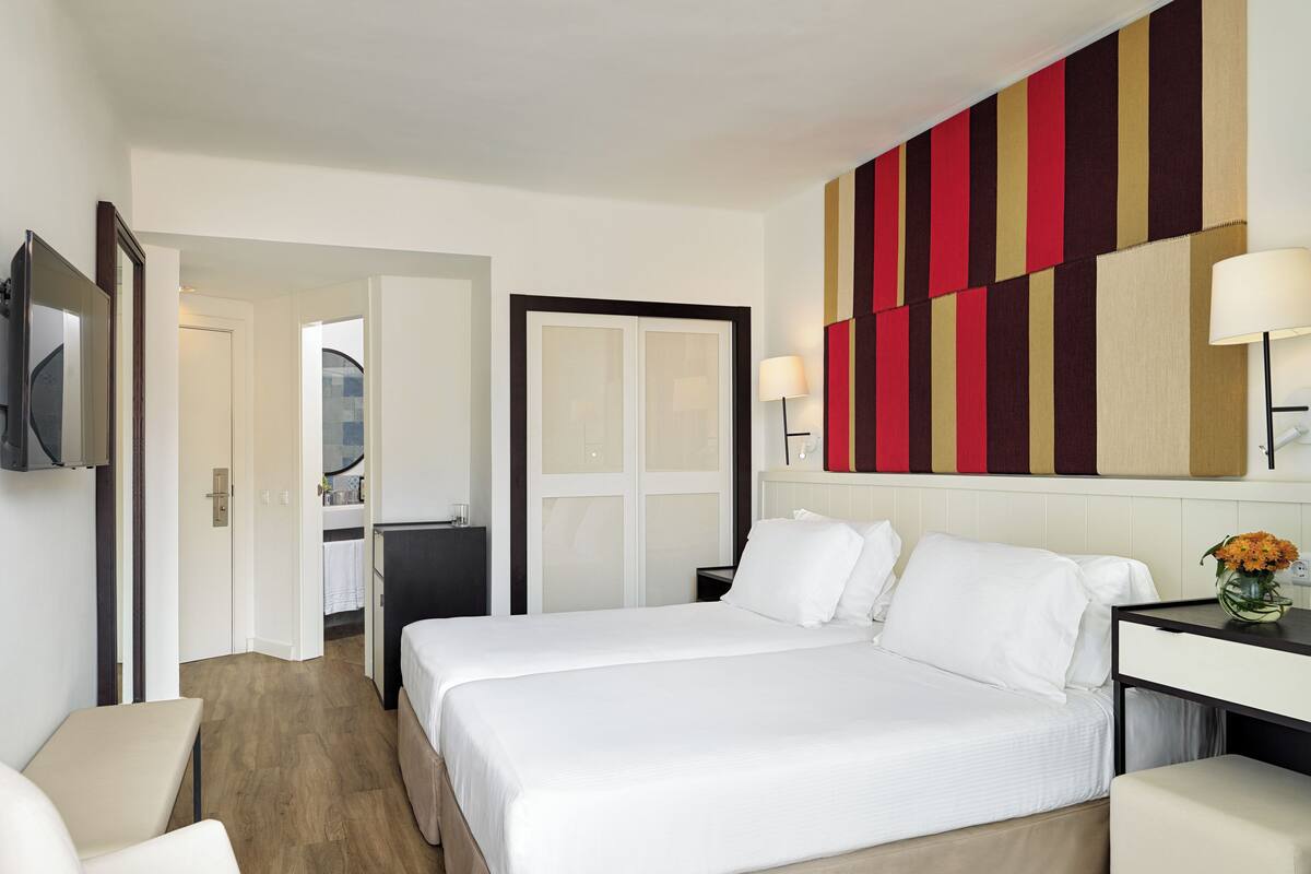 Quarto de hotel com uma cama grande, uma escrivaninha com candeeiro, uma televisão fixada na parede e uma casa de banho visível através de uma porta aberta.