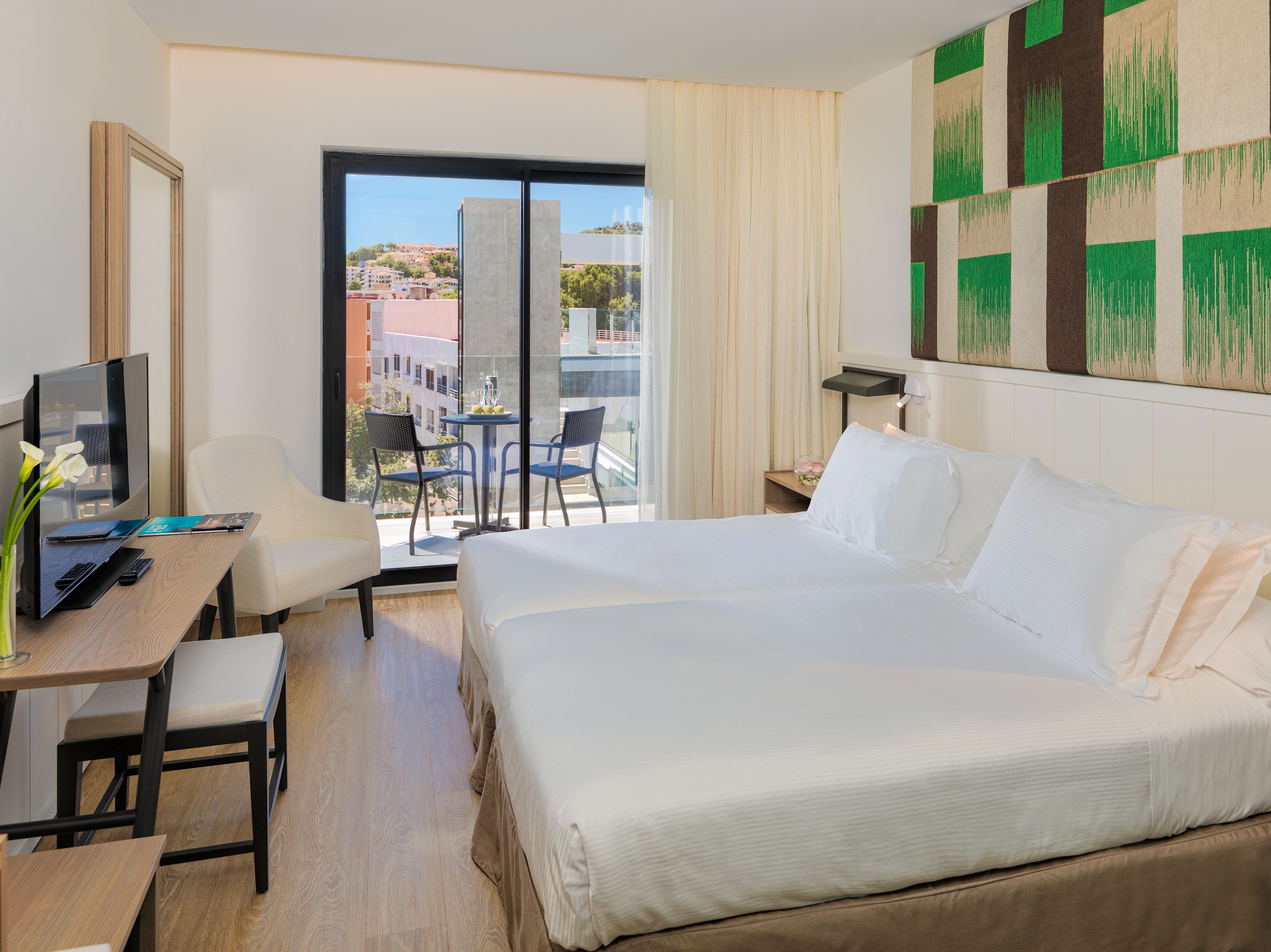 Quarto de hotel com uma cama, uma escrivaninha com computador, uma cadeira, uma mesinha com um vaso e vista para prédios através de uma grande janela.