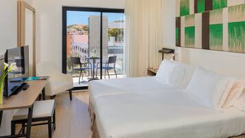 Quarto de hotel com uma cama, uma escrivaninha com computador, uma cadeira, uma mesinha com um vaso e vista para prédios através de uma grande janela.