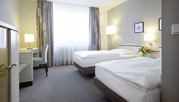 Quarto de hotel com duas camas, uma escrivaninha, uma cadeira e uma janela com cortinas.