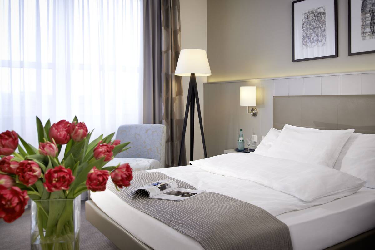 Quarto de hotel com uma cama bem arrumada, um vaso de tulipas vermelhas, um abajur e obras de arte emolduradas na parede.