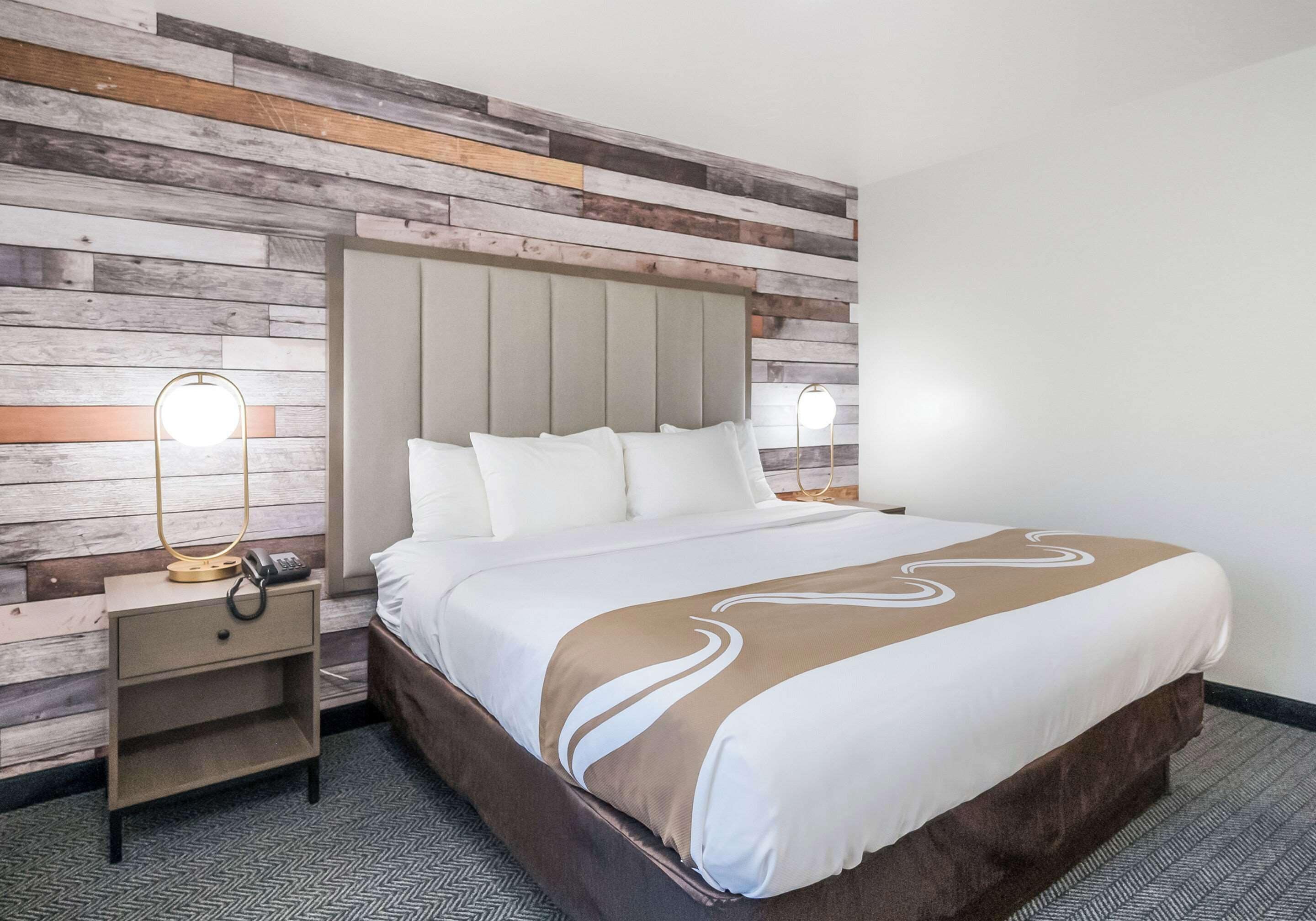 Quarto de hotel com cama, mesa de cabeceira, abajur e painel de parede de madeira.