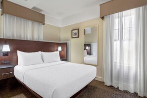 Quarto de hotel com uma cama grande, mesa de cabeceira com abajur, espelho e cortinas translúcidas.