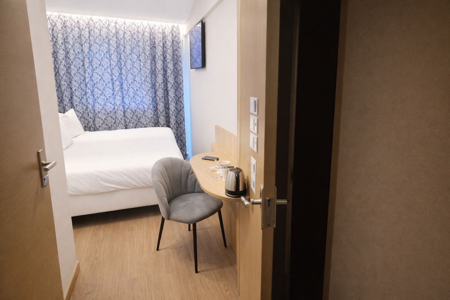 Quarto de hotel com cama, escrivaninha com cadeira, cafeteira e cortina estampada com flores.