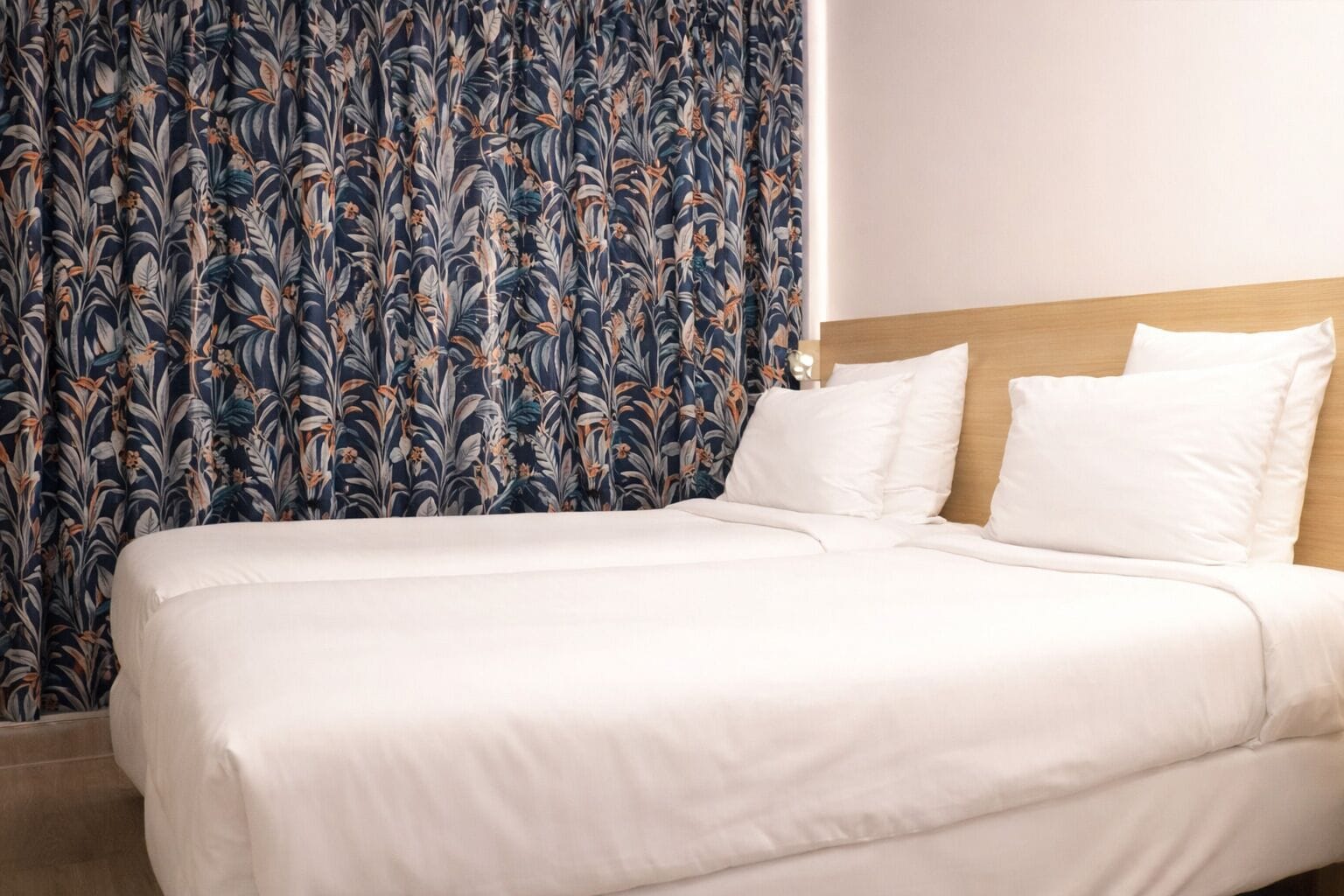 Quarto de hotel com cama, roupa de cama branca, cabeceira de madeira e cortina com padr ão floral.