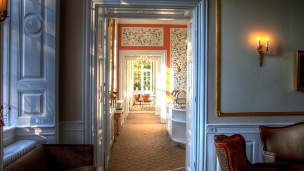 Um corredor com piso de carpete, uma área de jantar com cadeiras e uma mesa, e uma parede com papel de parede estampado com flores.