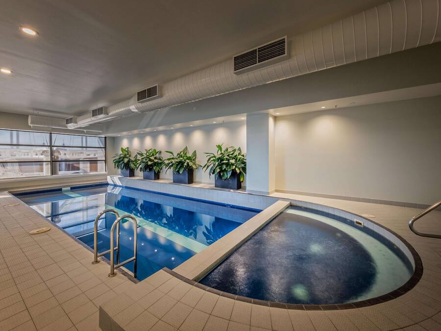 Uma piscina interior com vista desobstruída para a paisagem urbana através de amplas janelas, piso em azulejo e vários vasos de plantas ao longo da borda da piscina.