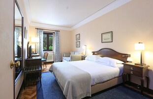 Quarto de hotel com uma cama grande, mesas de cabeceira, uma escrivaninha e uma cadeira. Há janelas com cortinas, uma porta e um espelho.