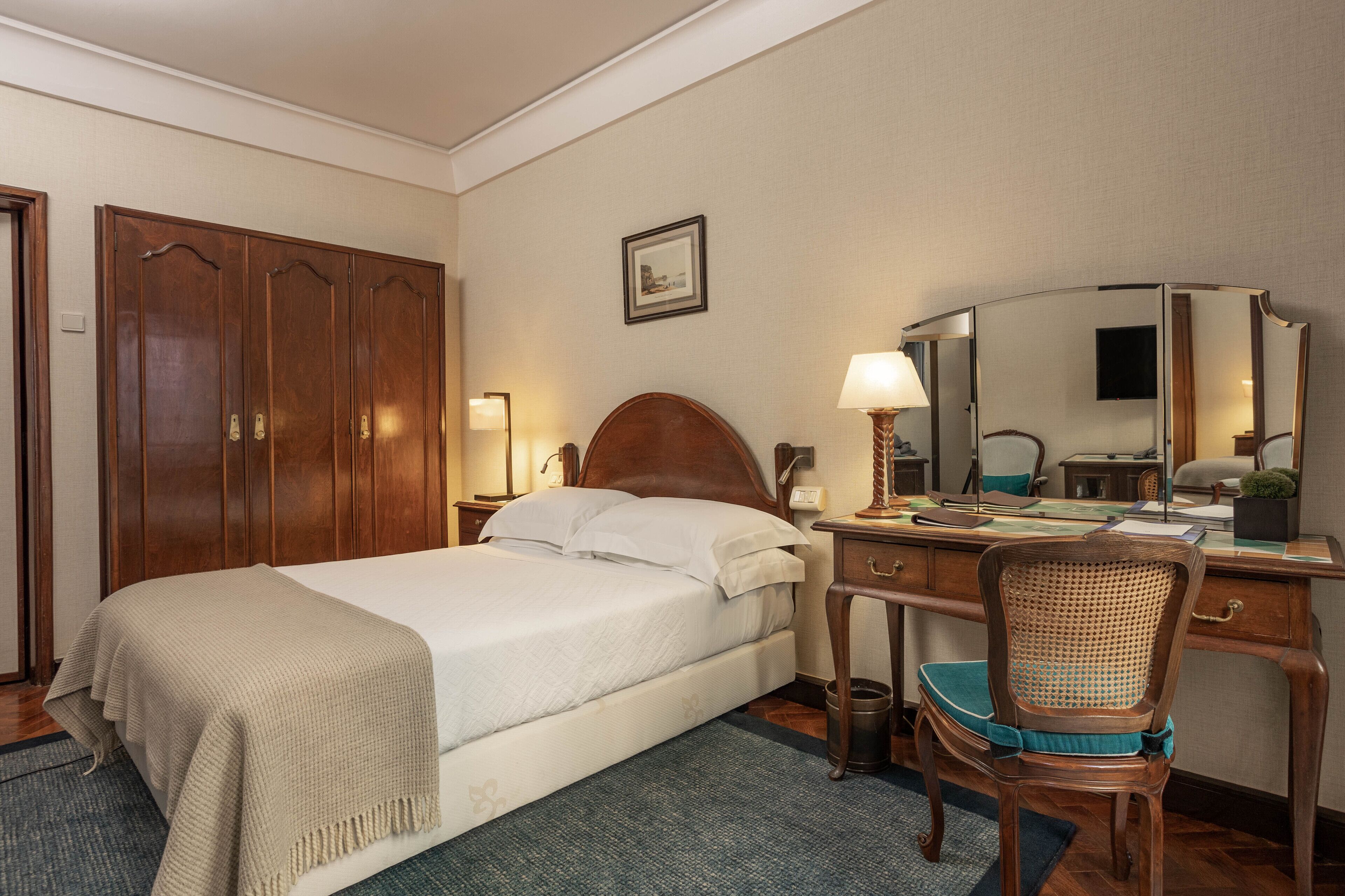 Quarto de hotel com cama, escrivaninha, cadeira, guarda-roupa e espelho.