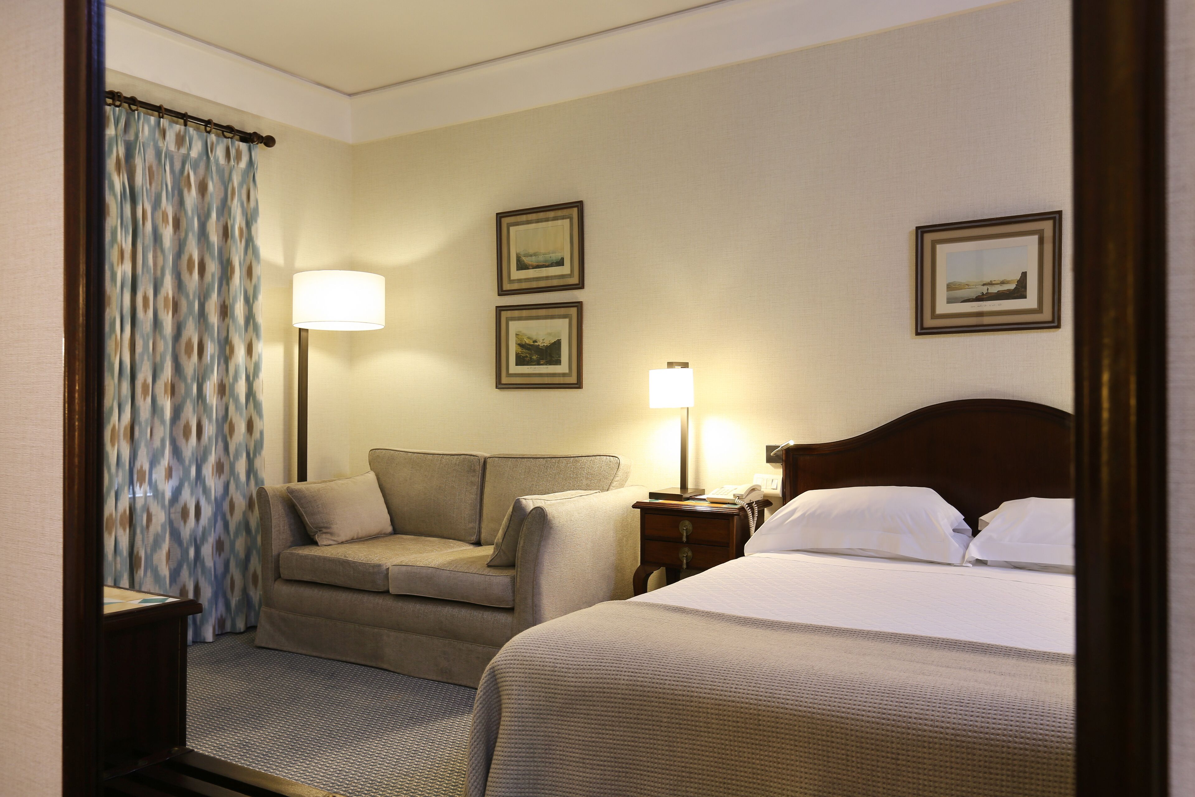 Quarto de hotel com cama, sofá, mesa de cabeceira, luminária e duas quadros na parede.