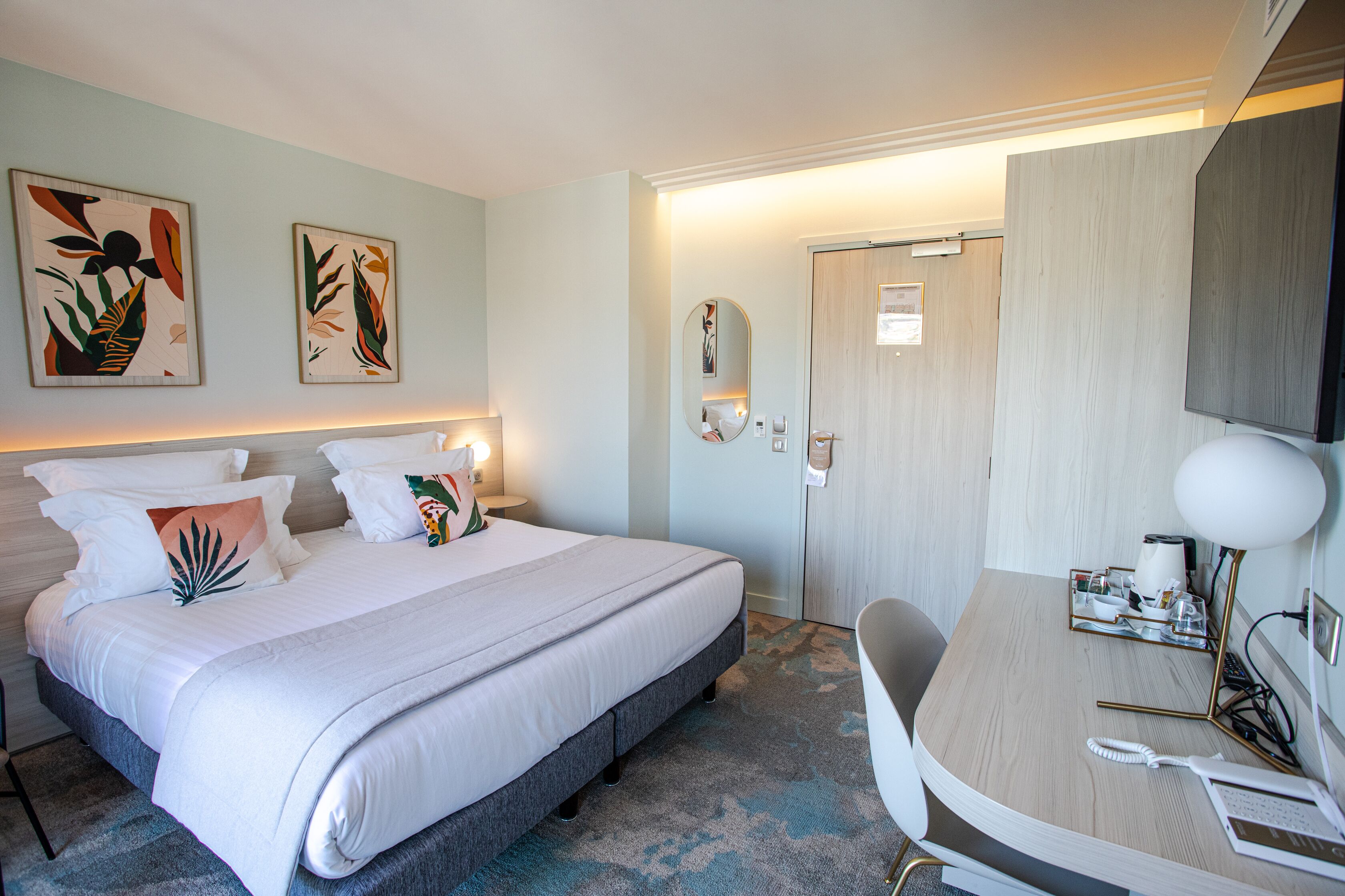 Quarto de hotel com cama, mesa de cabeceira, cadeira, escrivaninha e porta.