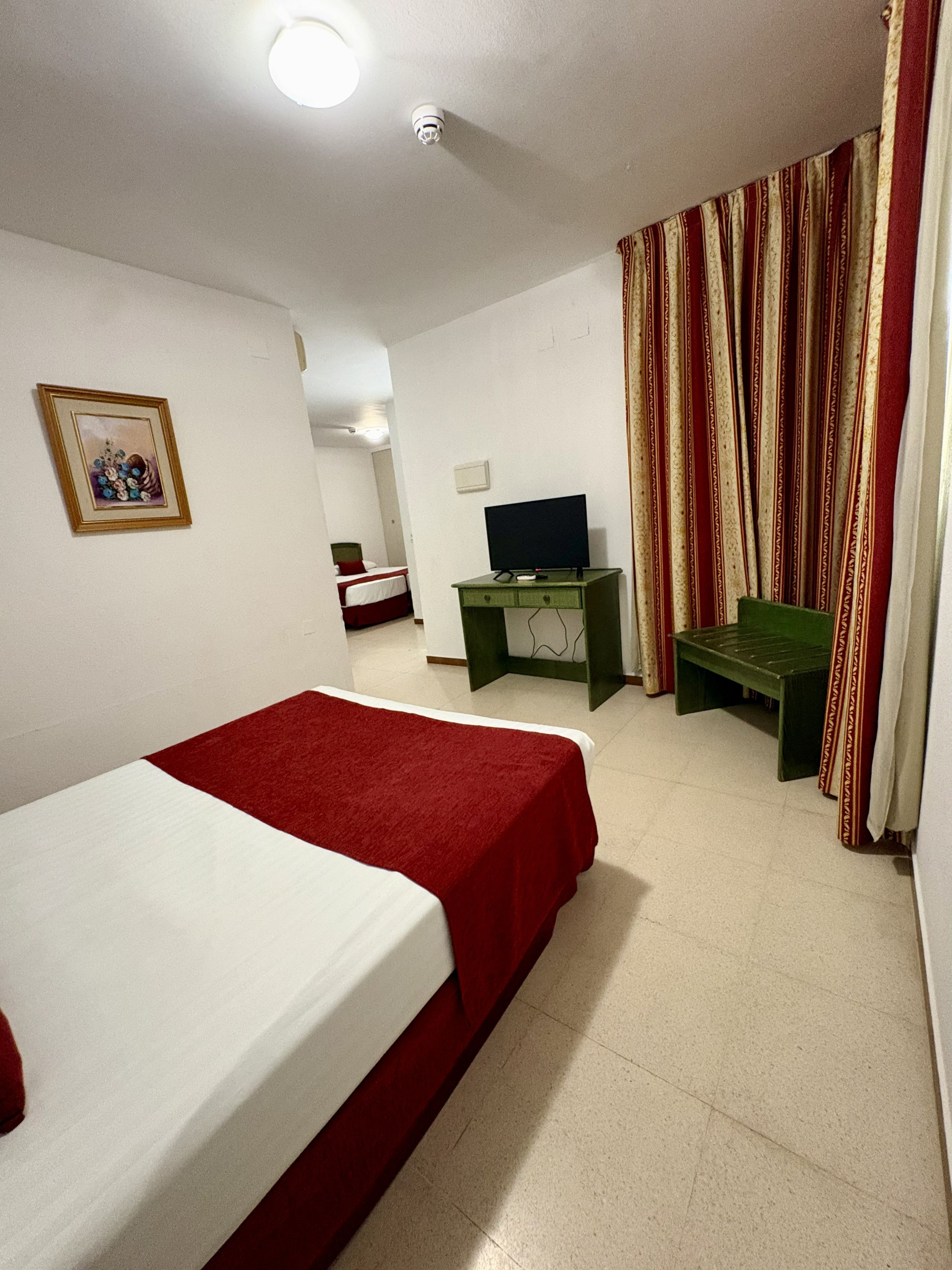 Quarto de hotel com cama, escrivaninha com computador, cadeira, televisão e janela com cortinas.