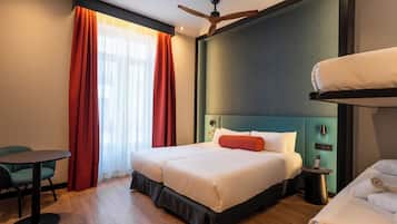 Quarto de hotel com cama, escrivaninha, cadeira, ventilador de teto e janela com cortinas.