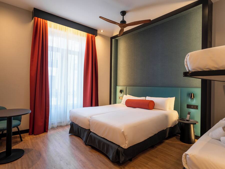 Quarto de hotel com cama, escrivaninha, cadeira, ventilador de teto e janela com cortinas.