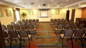 Sala de conferência com fileiras de cadeiras, uma tela de projetor e piso coberto por carpete.