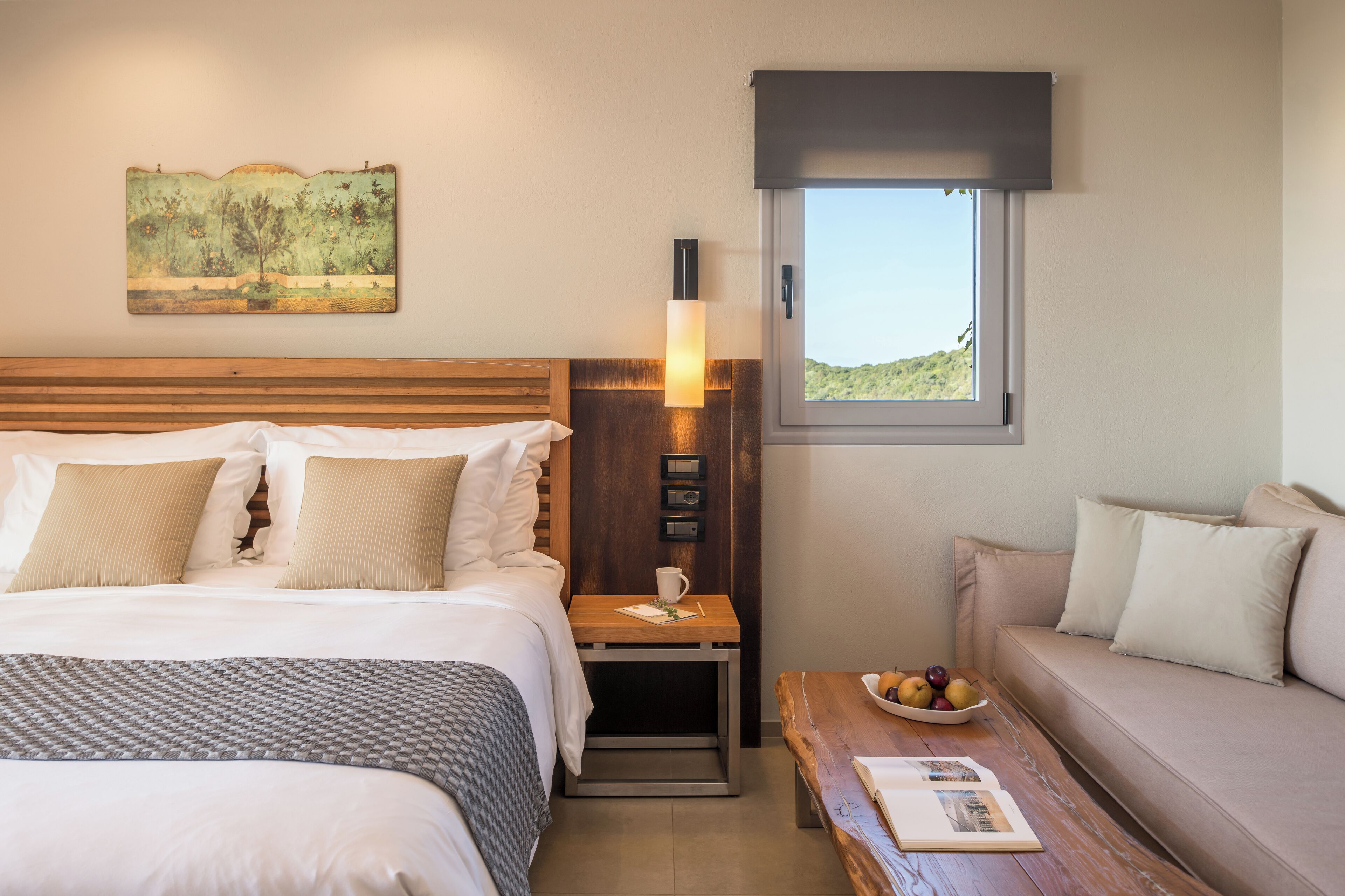 Quarto de hotel com uma cama grande, mesa de cabeceira com abajur, pequena mesa com frutas e um sofá.