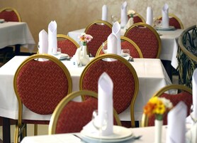 Uma área de refeições com mesas redondas, cadeiras estofadas vermelhas, toalhas de mesa brancas e guardanapos brancos.