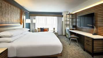 Quarto de hotel com uma cama grande, uma escrivaninha com televisão de tela plana, uma cadeira, uma mesinha e uma janela com cortinas.