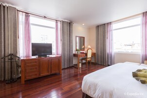 Quarto de hotel com secretária de madeira, televisão, cama e janelas amplas com cortinas.