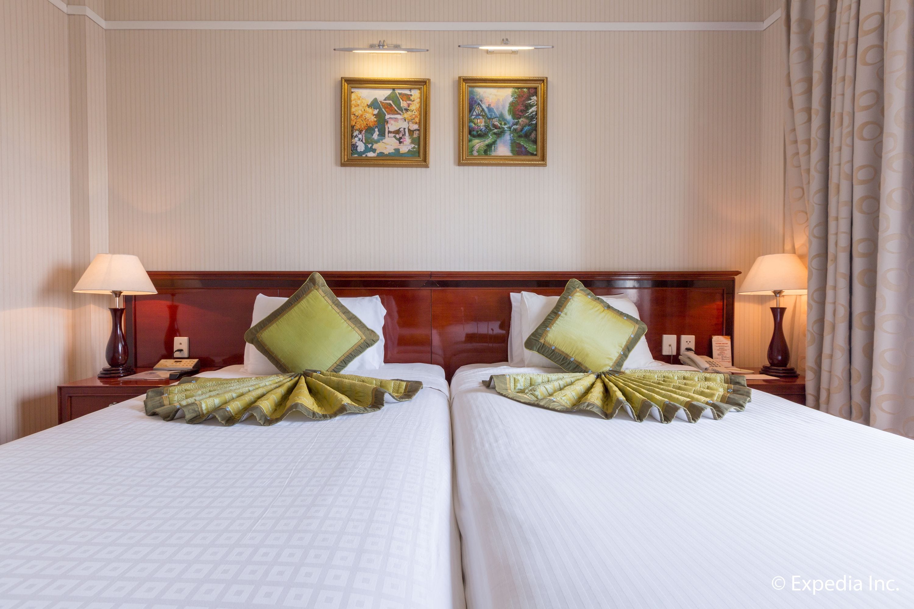 Quarto de hotel com duas camas, cada uma com cabeceira, abajures e almofadas decorativas. Existem duas pinturas emolduradas na parede acima das camas.