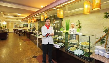 Uma área de buffet com diversos pratos expostos em vitrine, um funcionário em frente e uma área de estar ao fundo.